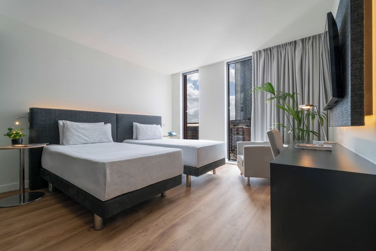 Quarto de hotel moderno com duas camas, uma escrivaninha, uma cadeira e uma janela grande com cortinas.