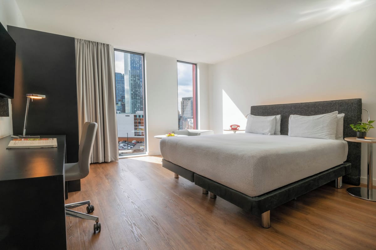 Quarto de hotel moderno com uma cama grande, uma escrivaninha com cadeira, uma pequena mesa com uma planta e vista para a cidade através de grandes janelas.