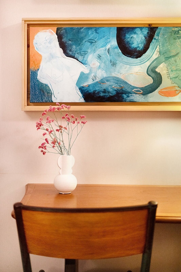 Pintura abstrata emoldurada numa parede, acima de uma mesa de madeira com um vaso branco contendo flores cor-de-rosa.