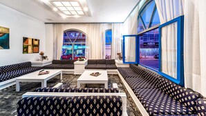 Um hall de hotel moderno com sofás estampados, uma mesa de centro branca e grandes janelas com vista para a rua.