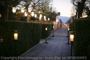 Uma trilha pavimentada, ladeada por sebes e iluminada por postes de rua, conduz a um edifício com uma placa vermelha e branca.