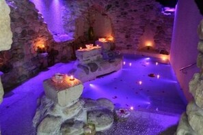 Quarto de paredes de pedra com uma piscina central, velas e um banco de pedra.