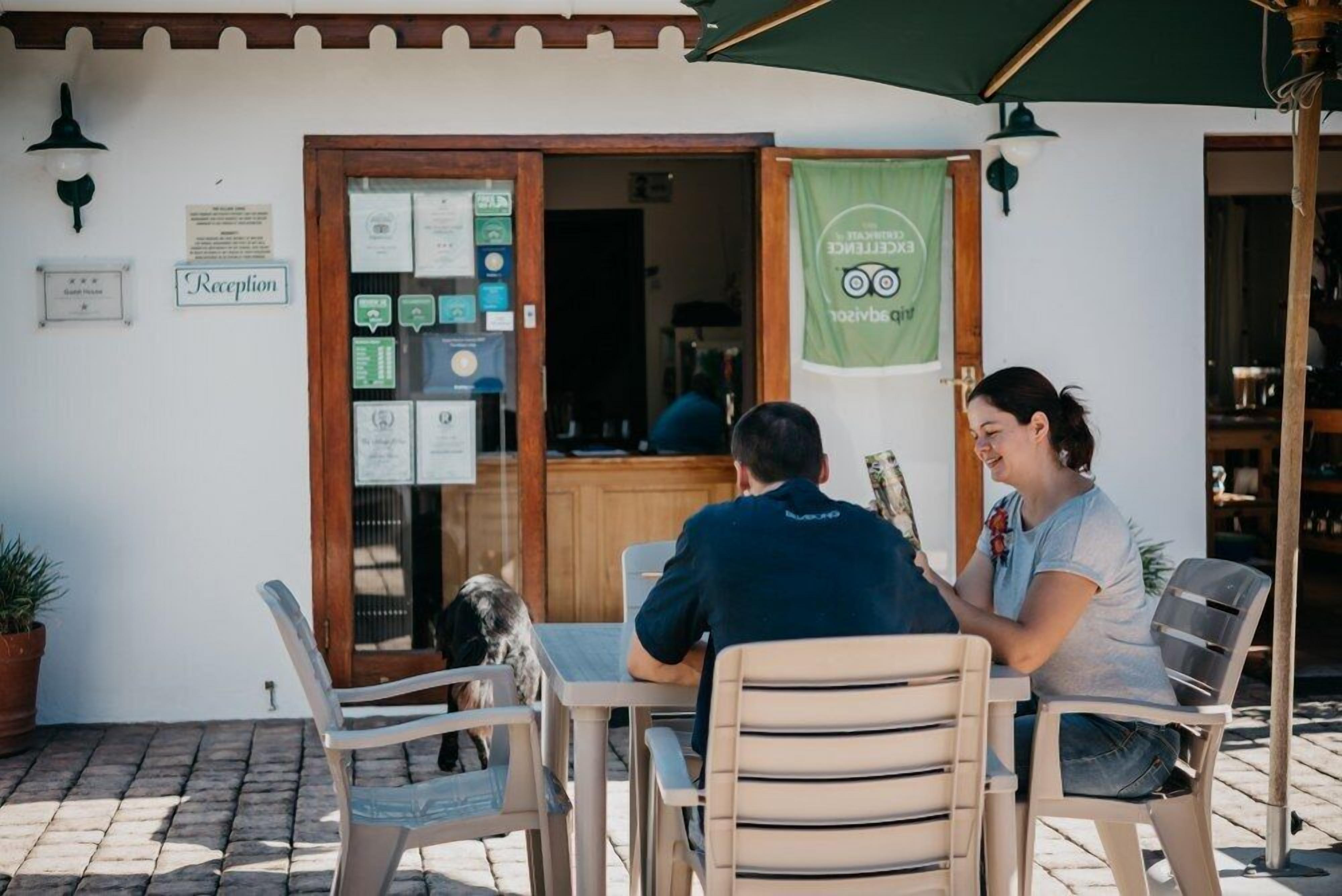 Uma área externa com mesas e cadeiras, uma mesa de recepção, um cão e um banner promovendo um negócio local.