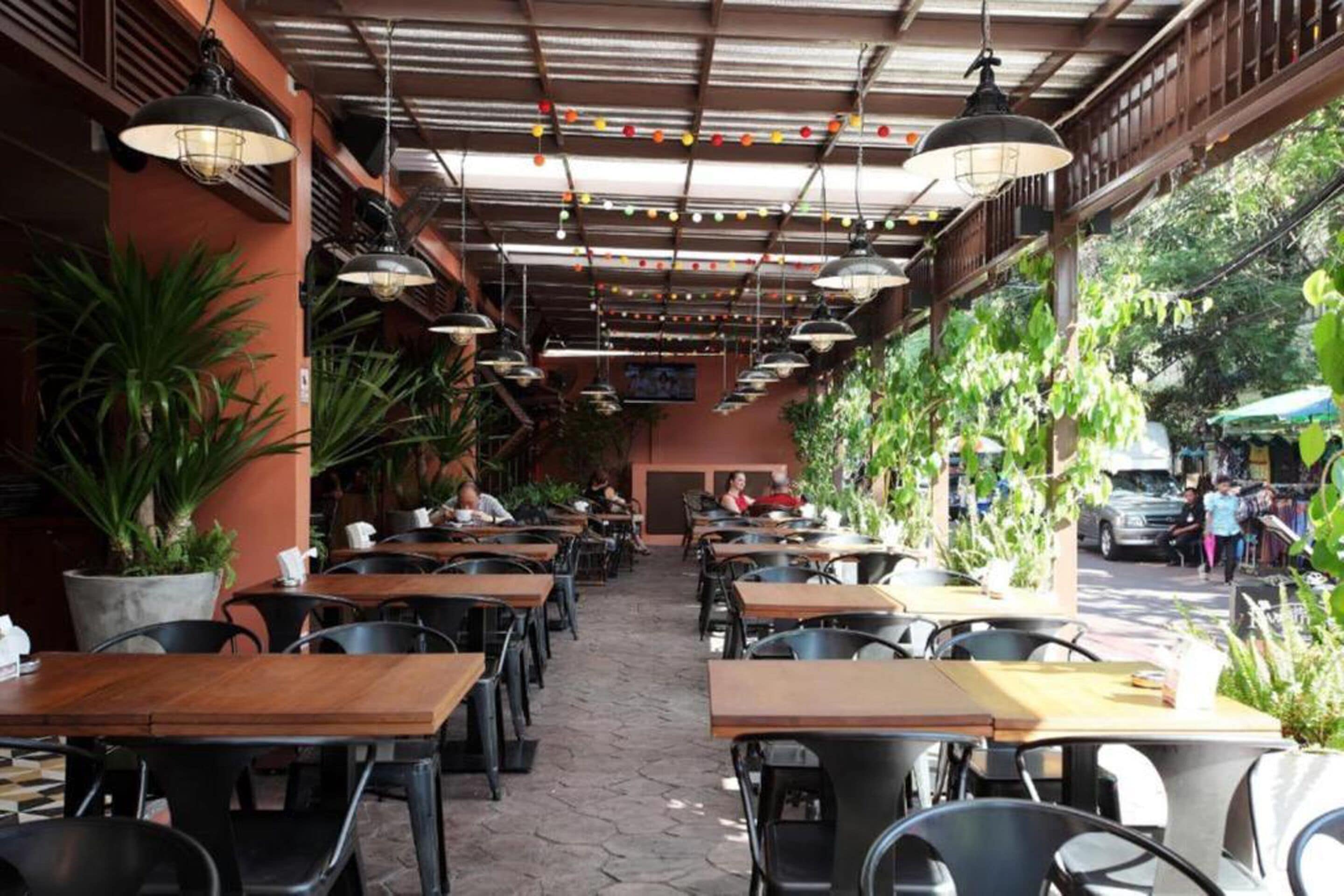 Café ao ar livre com mesas, cadeiras, plantas em vasos e luzes penduradas.