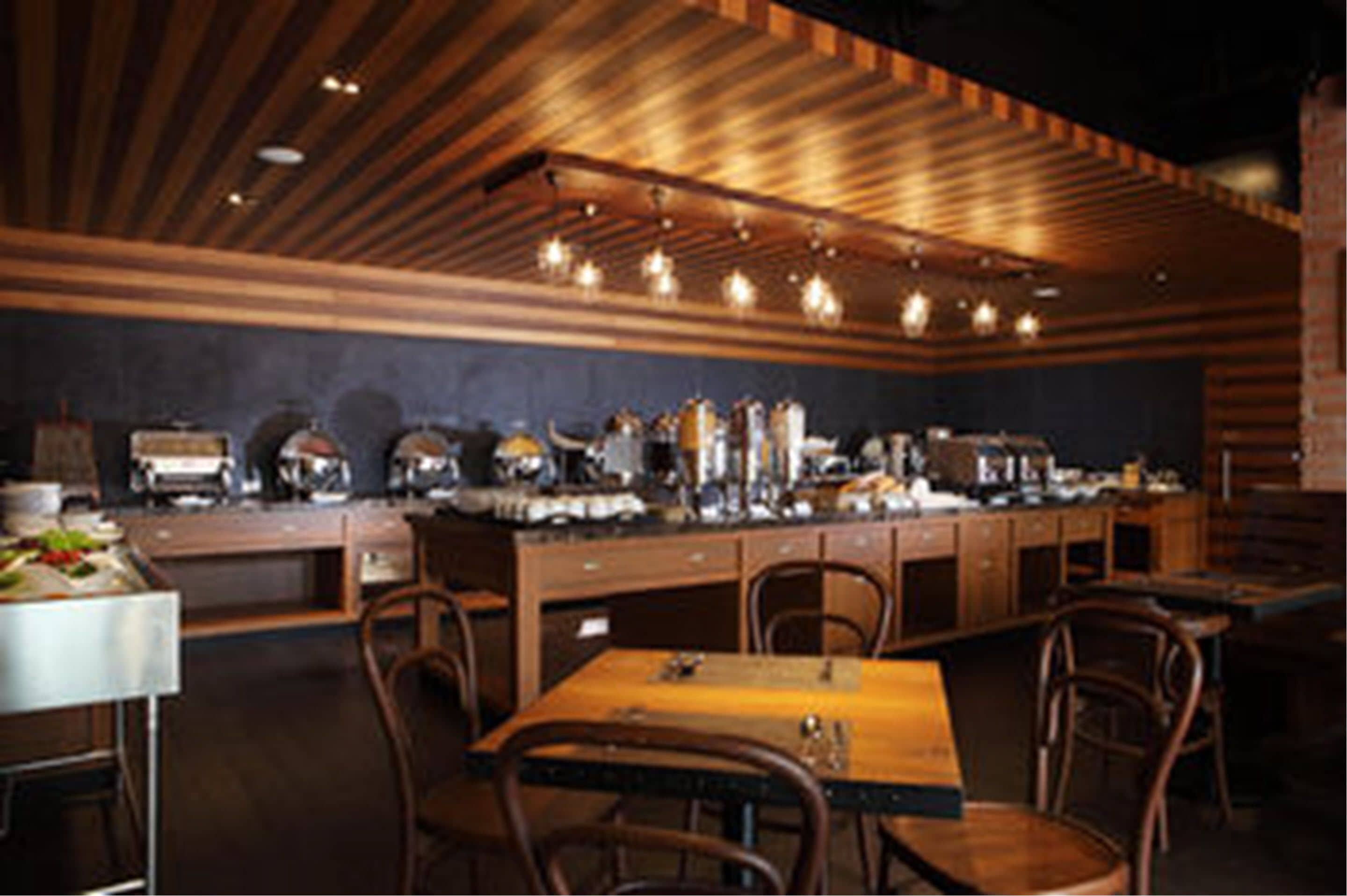 Restaurante com teto de madeira e um balcão comprido com diversos equipamentos de cozinha. Várias mesas e cadeiras de madeira estão dispostas.