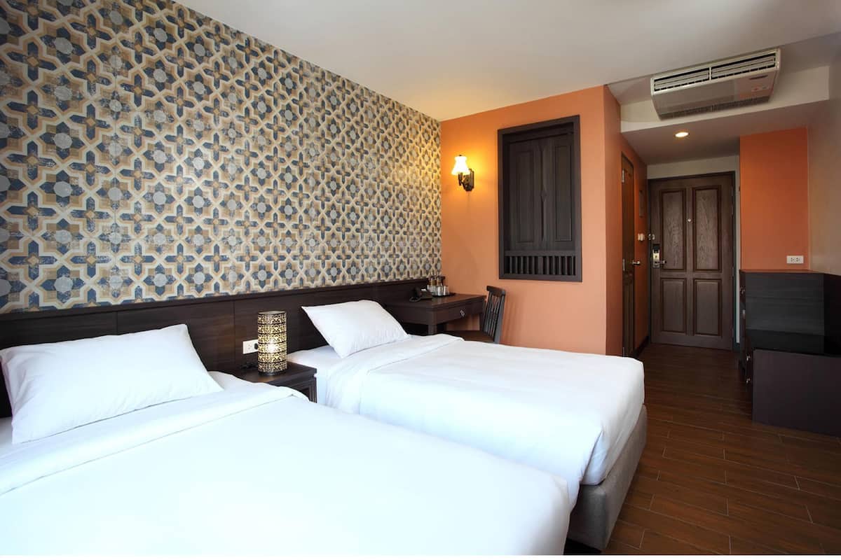 Quarto de hotel com duas camas, parede estampada, mesa de cabeceira com abajur, televisão e aparelho de ar condicionado.