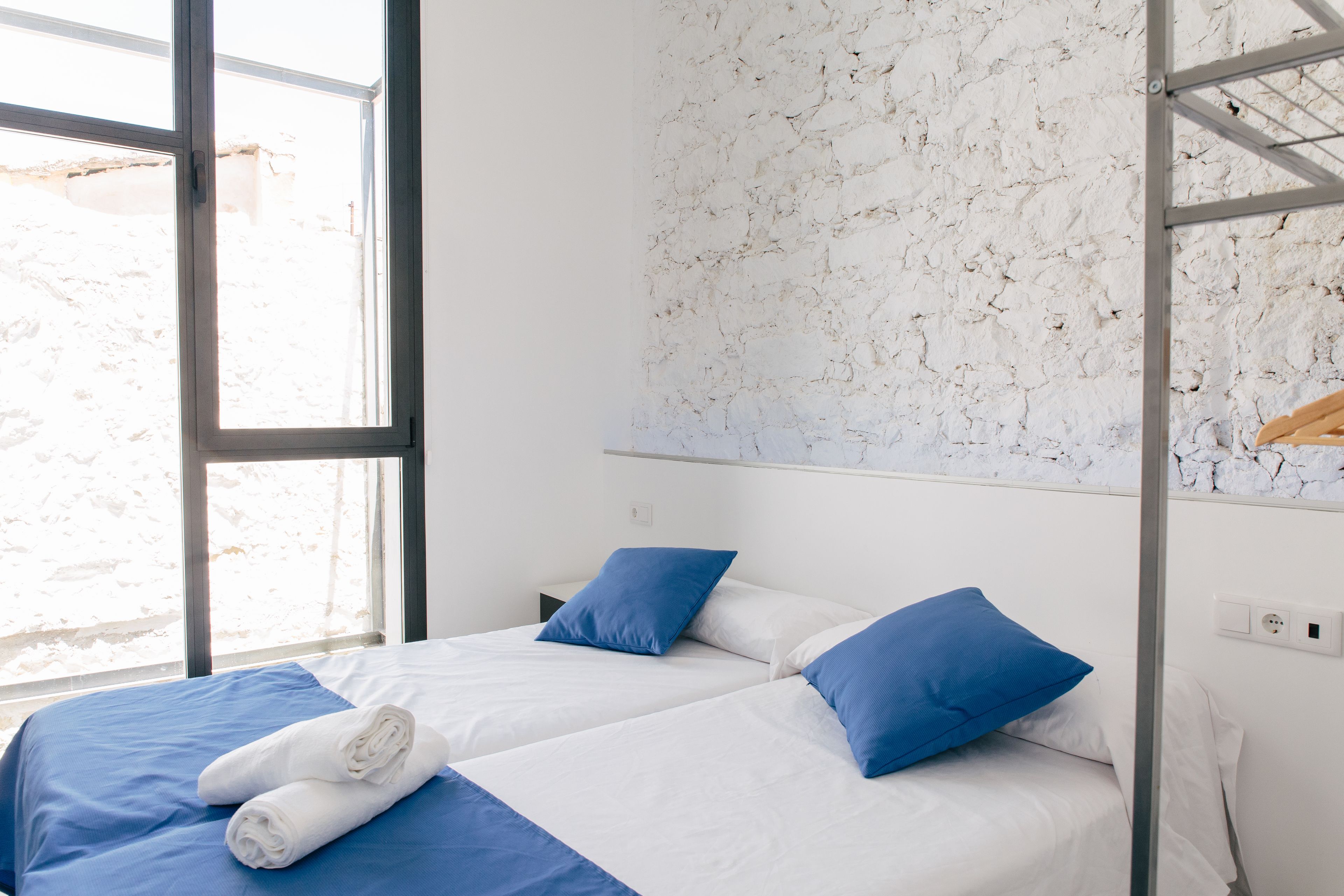 Quarto de hotel com uma cama, duas almofadas, uma toalha enrolada, uma janela e uma parede com textura.