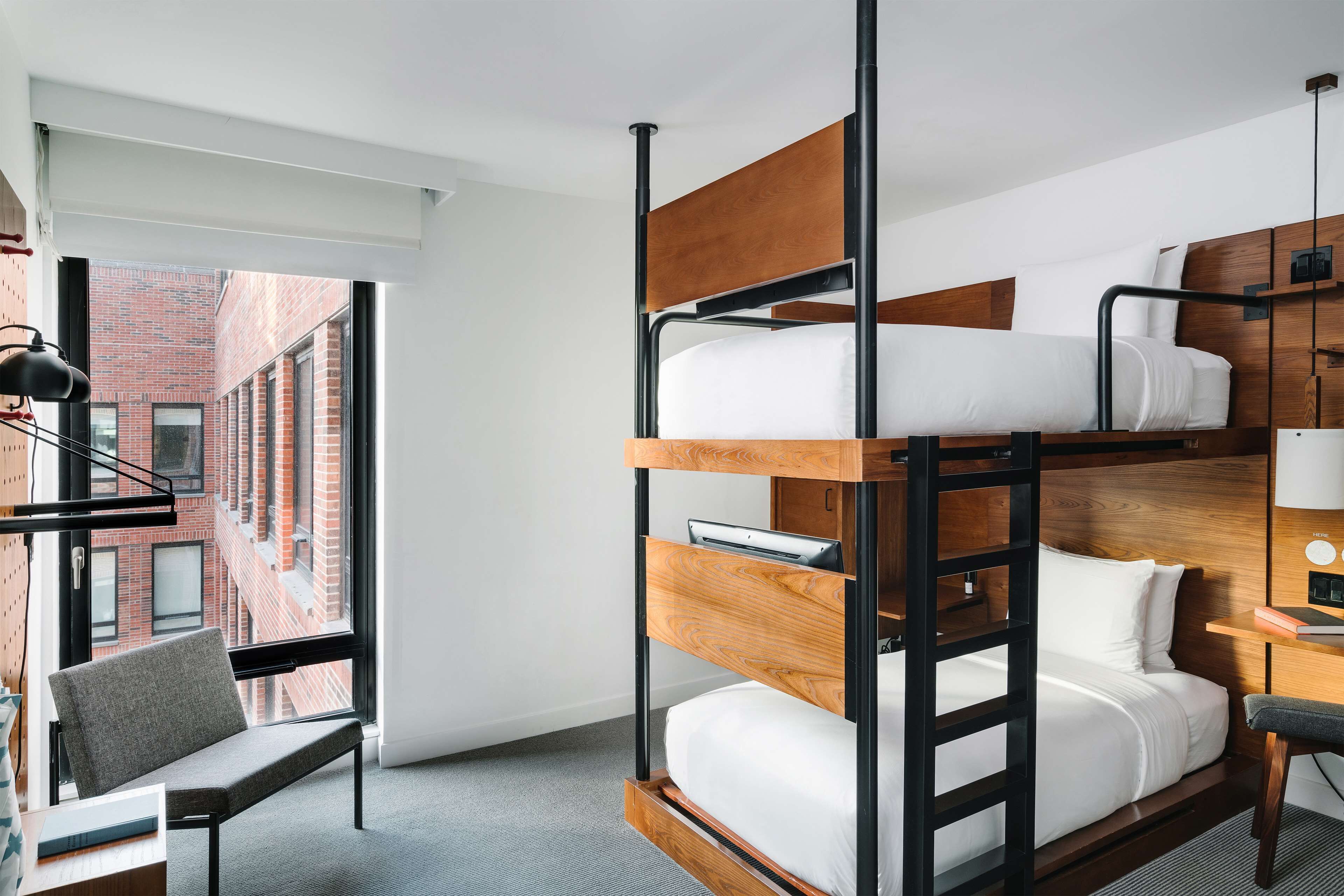 Uma cama beliche com cabeceira de madeira e estrutura de metal preta, uma escrivaninha com uma cadeira e vista de um edifício de tijolos por uma janela grande.