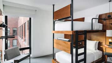 Uma cama beliche com cabeceira de madeira e estrutura de metal preta, uma escrivaninha com uma cadeira e vista de um edifício de tijolos por uma janela grande.