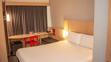 Quarto de hotel com uma cama, uma escrivaninha, uma cadeira e uma mesinha.