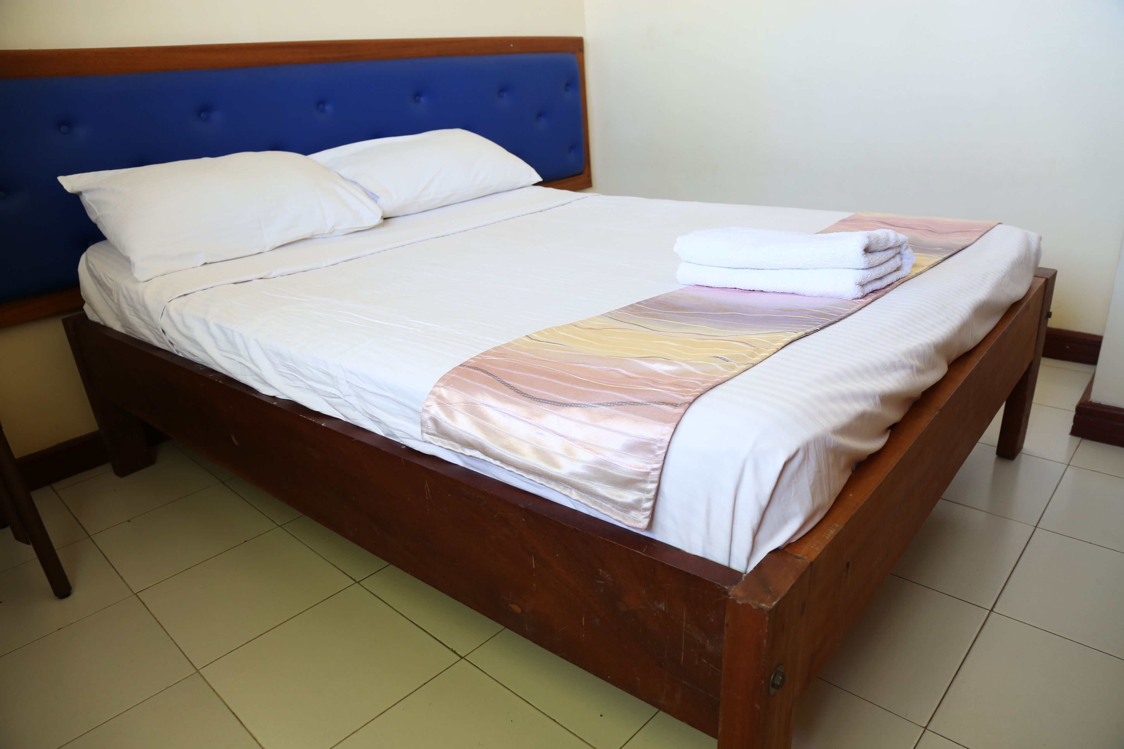 Cama bem arrumada com lençóis brancos e toalha dobrada, cabeceira estofada azul e estrutura de cama de madeira.