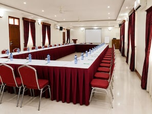 Uma sala de conferências com uma mesa comprida, cadeiras vermelhas e garrafas de água.
