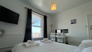 Quarto de hotel com uma cama, uma secretária com cadeira, televisão e janela com cortinas.