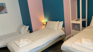 Quarto com duas camas de solteiro, uma mesa de cabeceira de madeira com abajur e prateleira com ventilador.