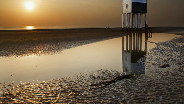 Um farol sobre pilares na praia, com o sol se pondo ao fundo.