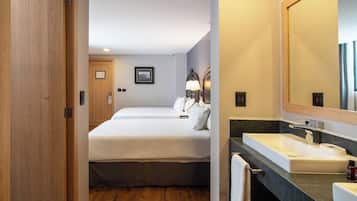 Quarto de hotel com porta de madeira, cama com roupa de cama branca, mesa de cabeceira com abajur, banheiro com pia e espelho e um quadro na parede.