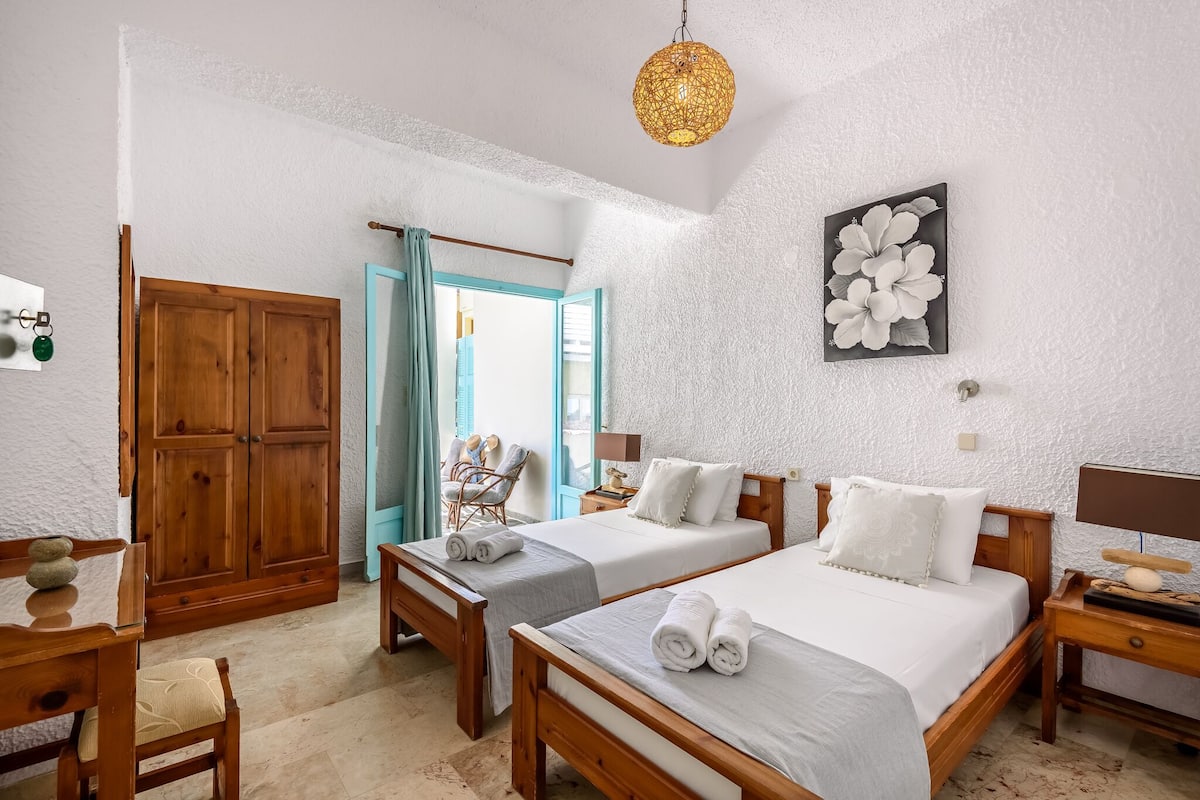 Quarto de hotel com duas camas de madeira, uma mesa de jantar com cadeiras, um guarda-roupa e uma janela grande com cortinas.