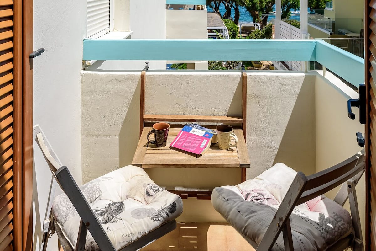 Varanda com vista para o mar, uma cadeira de madeira, uma mesa pequena com um livro e uma caneca, e um prédio branco com persianas.
