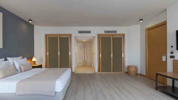 Quarto de hotel moderno com uma cama grande, uma mesa de cabeceira, uma luminária e um banco de madeira. Há dois armários e uma porta que dá para um corredor.
