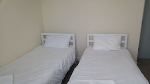 Duas camas de solteiro com cabeceiras e almofadas brancas em um quarto com paredes lisas.