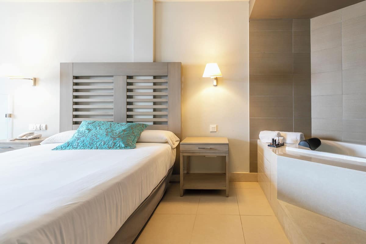 Quarto de hotel moderno com cama, mesa de cabeceira, luminária e banheira.