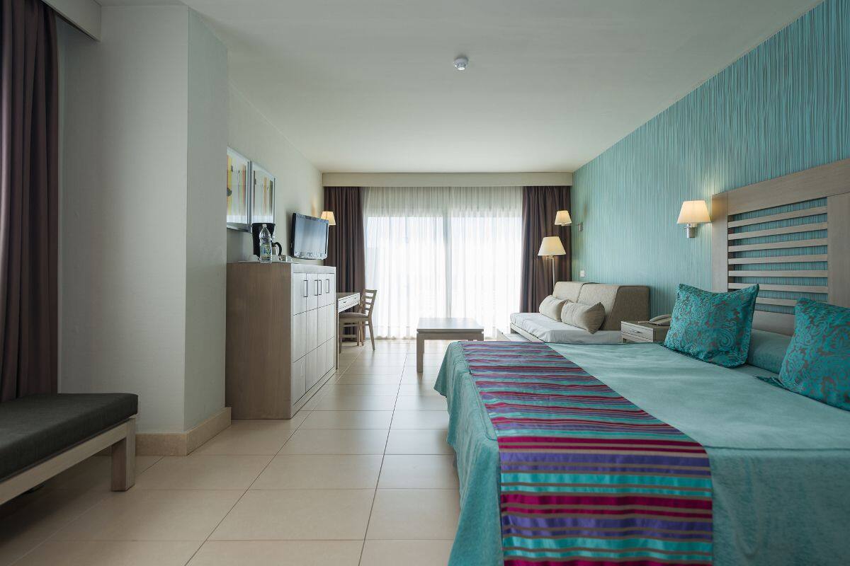 Quarto de hotel com cama, secretária, cadeira, televisão e minibar.