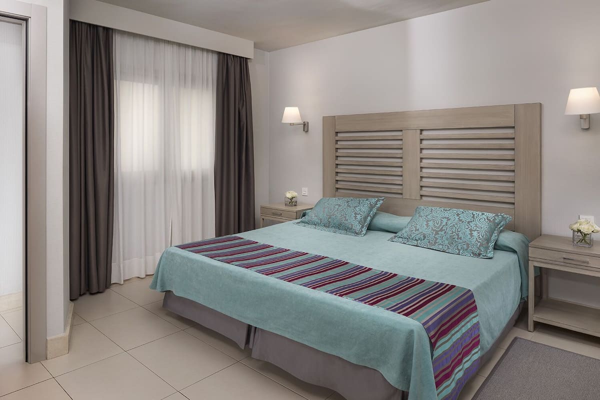 Quarto de hotel com uma cama, duas mesas de cabeceira, uma luminária e uma janela com cortinas.