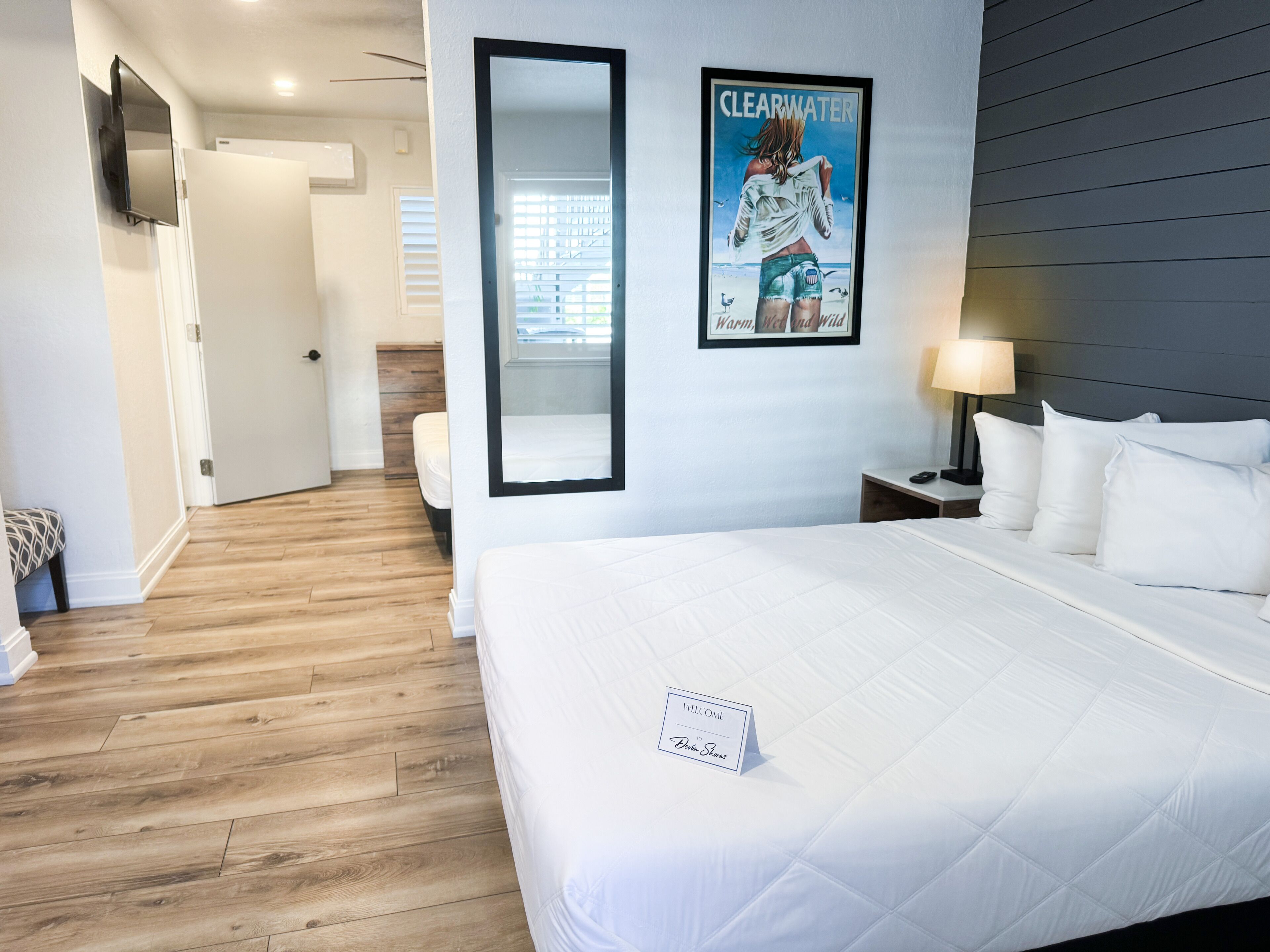 Quarto de hotel com uma cama grande, uma mesa de cabeceira com abajur, um quadro, um espelho e piso de madeira.