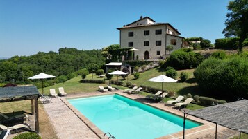 Uma piscina com espreguiçadeiras e guarda-sóis, cercada por vegetação e um prédio grande ao fundo.