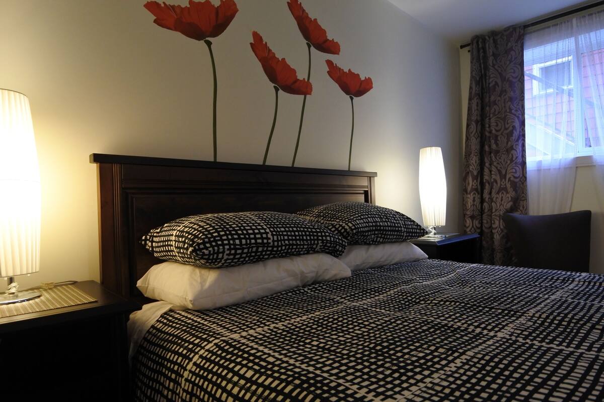 Cama bem arrumada com roupa de cama estampada, mesa de cabeceira com abajur e parede decorada com flores vermelhas.