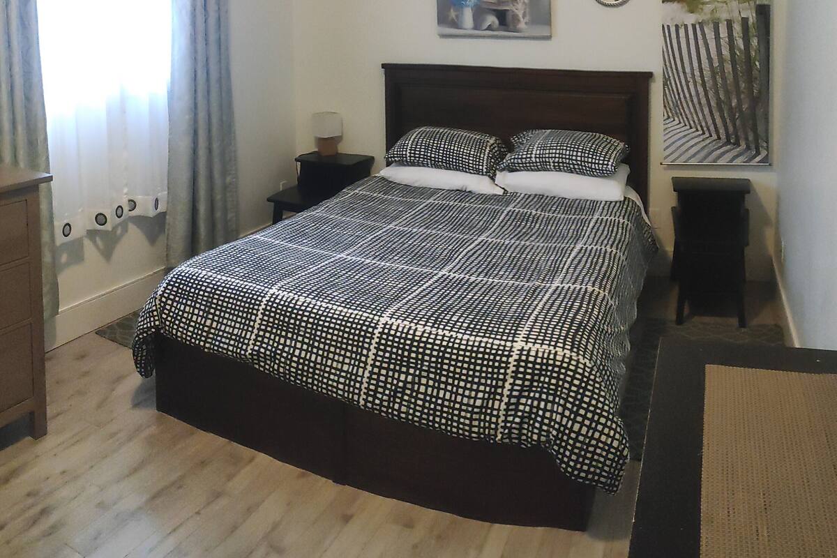 Quarto com cama grande, mesa de cabeceira, cômoda, pintura e janela com cortinas.