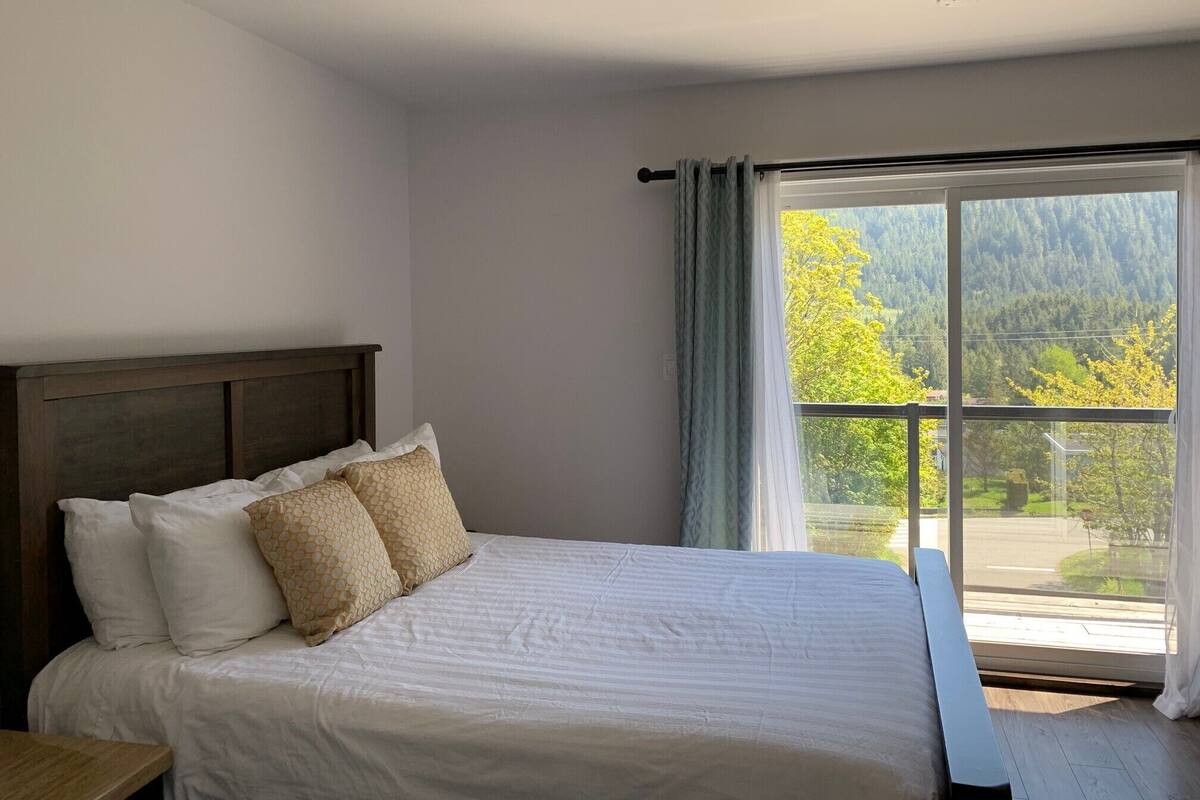 Quarto com uma cama grande, cabeceira de madeira e vista para uma paisagem verde através de uma janela ampla.