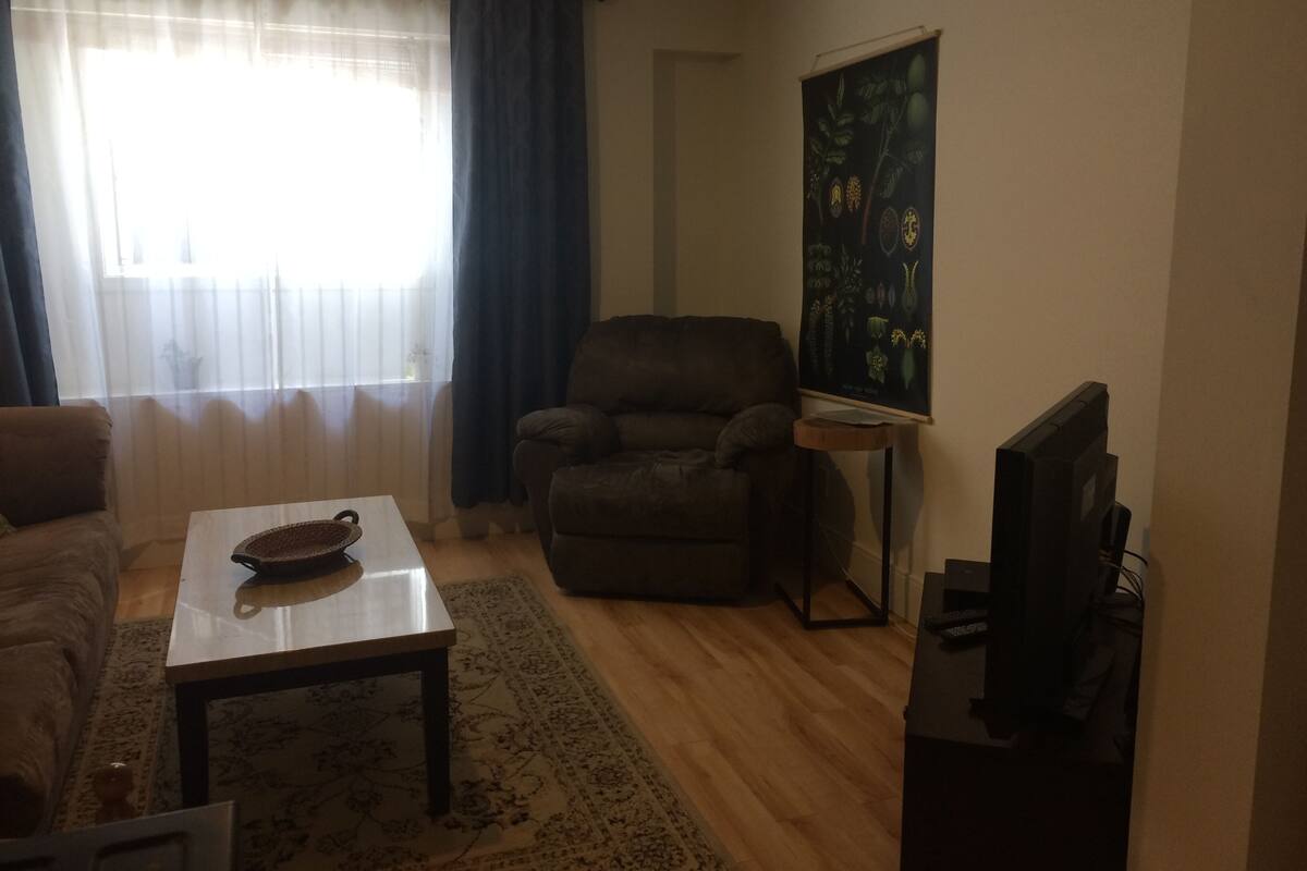 Uma sala de estar com sofá, mesa de centro, poltrona reclinável, televisão e um pôster na parede.