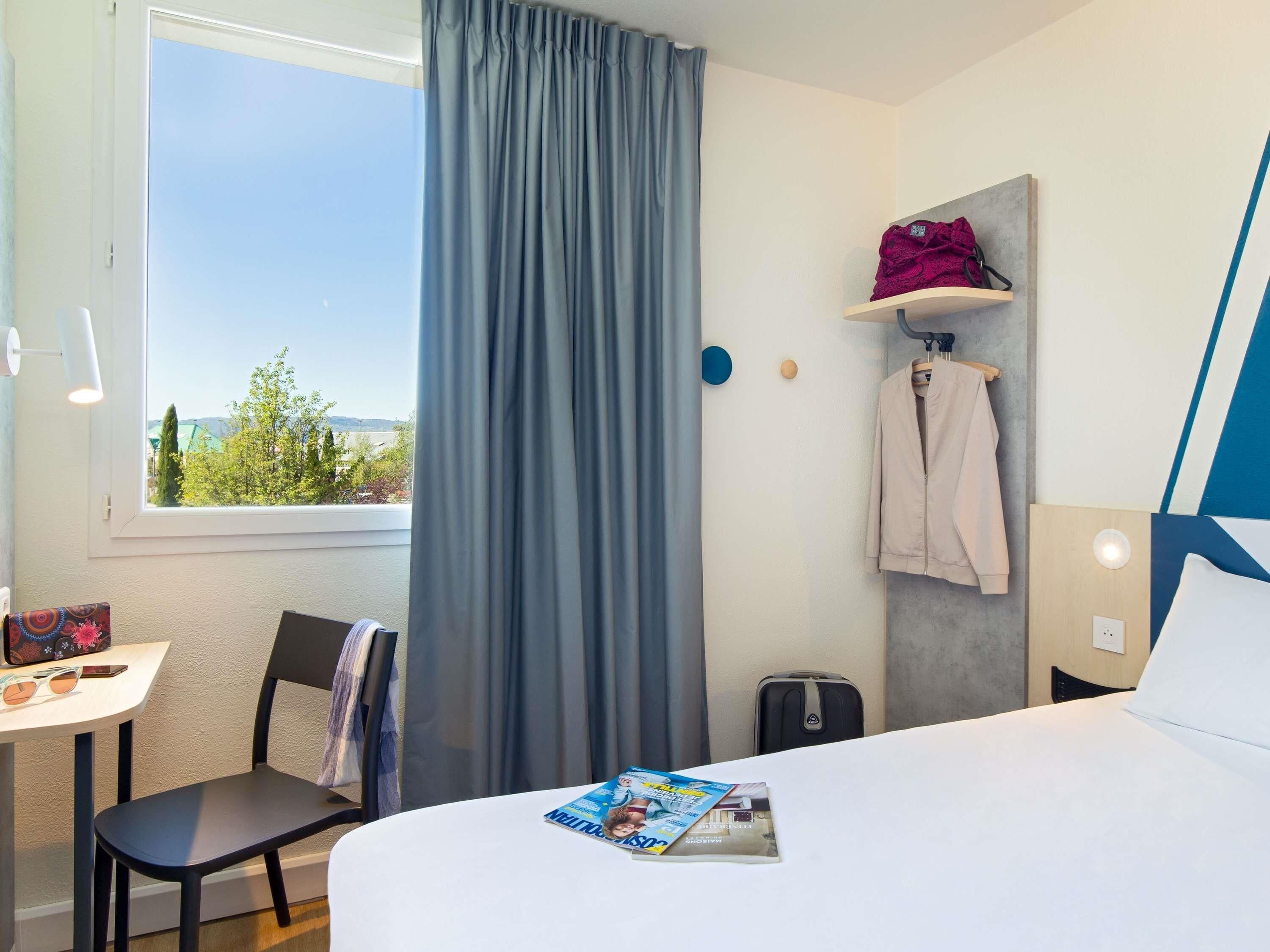 Quarto de hotel com cama, escrivaninha com cadeira, janela com cortinas, prateleira na parede com um casaco e uma revista sobre a cama.