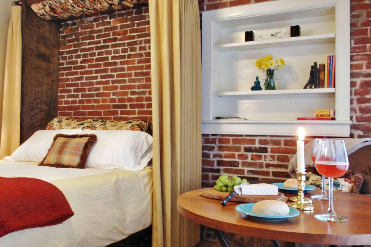 Quarto de hotel com parede de tijolos, cama com roupa branca, abajur, mesa pequena com um prato de comida, garrafa de vinho e uma vela.