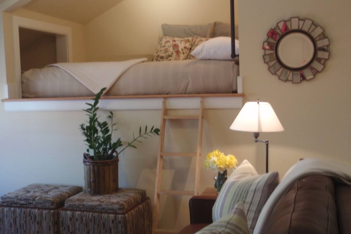 Quarto no estilo loft com cama beliche, uma escada, mesa de cabeceira com abajur, planta em vaso e um banquinho.