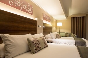 Quarto de hotel com duas camas, uma cabeceira de madeira, uma luminária na parede e uma grande janela com cortinas.