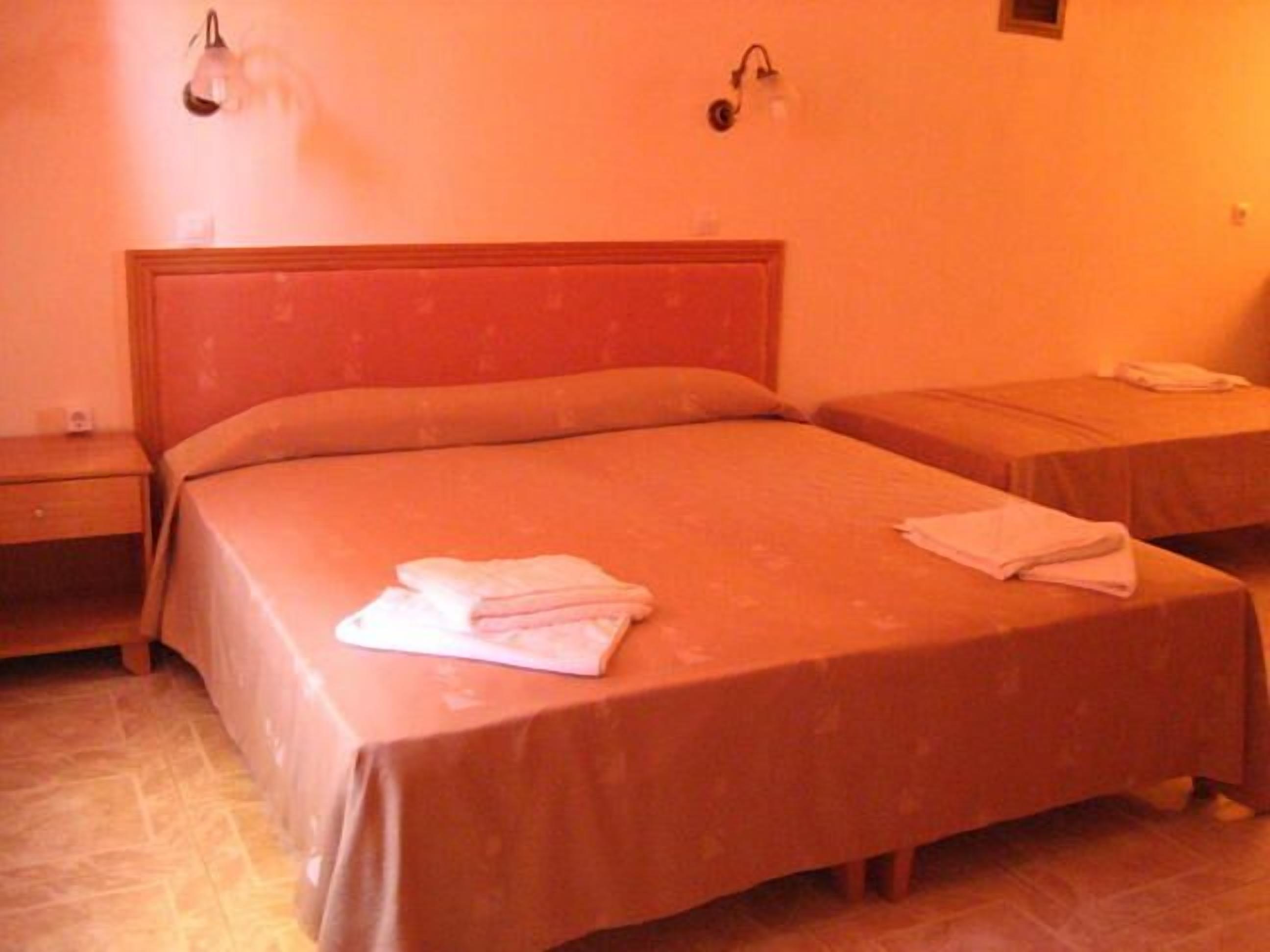 Quarto de hotel com uma cama grande, uma cama menor, uma mesinha de cabeceira e duas luminárias fixadas na parede.
