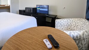 Quarto de hotel com uma cama, uma mesa redonda de madeira com um controle remoto, uma poltrona estampada, uma escrivaninha preta com um micro-ondas e uma televisão exibindo um menu de configuração.