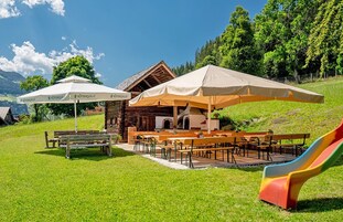 Um chalé rústico com área externa para sentar e um escorrega, cercado por vegetação exuberante e montanhas.