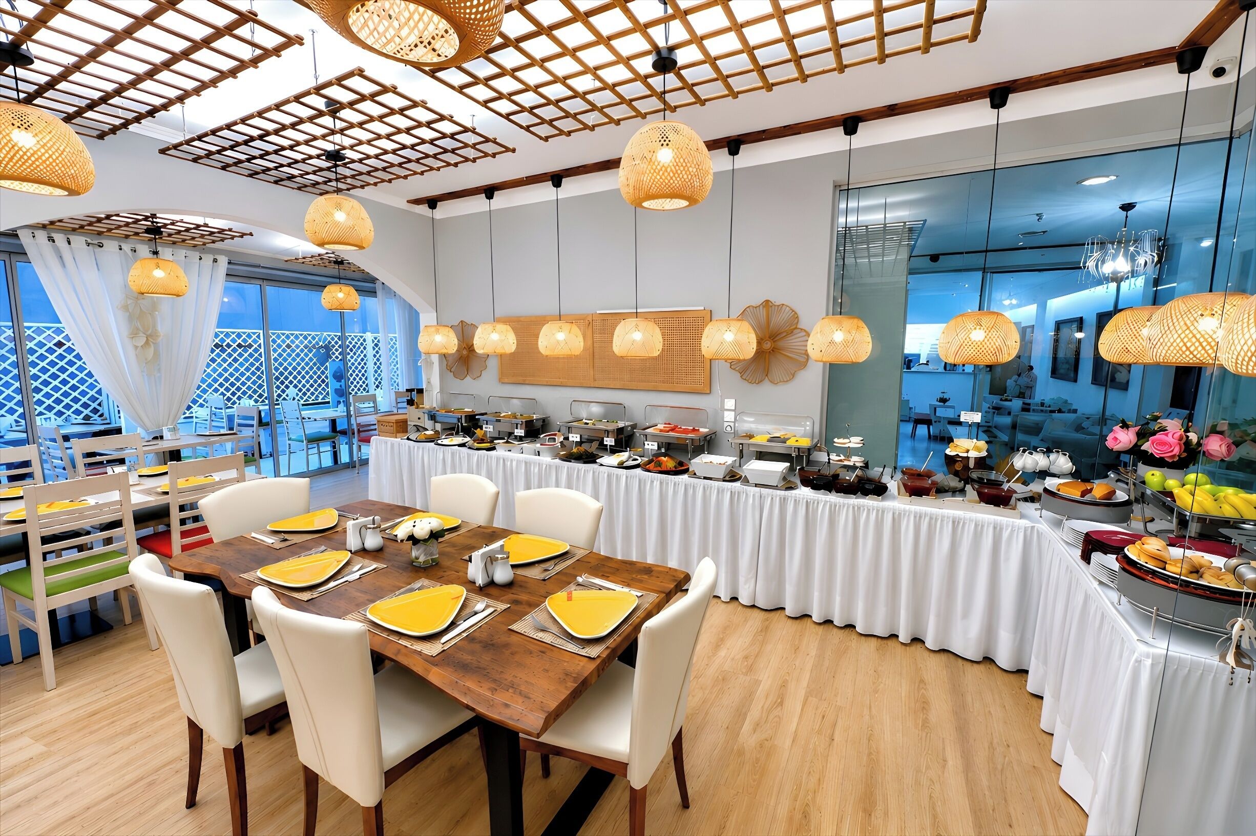 Uma área de refeições com mesas preparadas para uma refeição, um buffet com diversos alimentos e vista para o exterior através de grandes janelas.