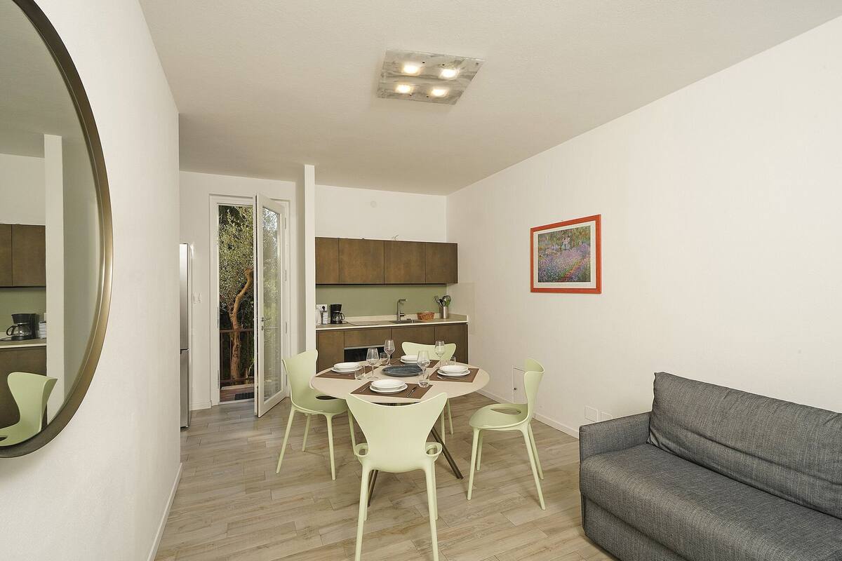 Uma sala de estar moderna com um sofá cinzento, uma mesa de jantar com cadeiras, uma cozinha com armários e um quadro na parede.