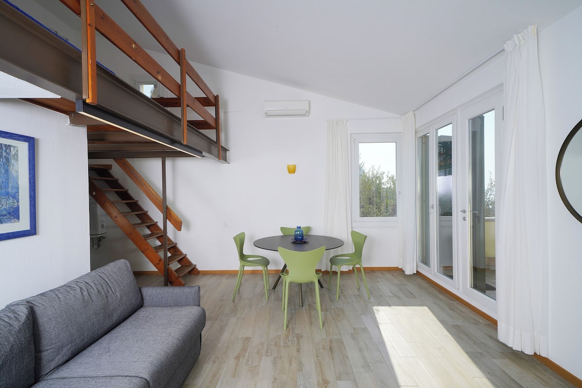Um quarto com uma escada, uma mesa de jantar com cadeiras, um sofá e uma janela grande com cortinas.