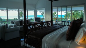 Quarto com cama grande, escrivaninha e cadeira, com vista para uma praia e palmeiras.