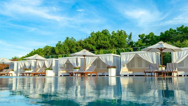 Área de piscina com várias espreguiçadeiras e guarda-sóis, rodeada por toldos brancos e vegetação.