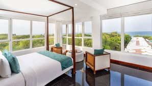 Quarto de hotel moderno com uma cama grande, área de estar com uma poltrona e uma mesa pequena, e vista para o mar e vegetação.