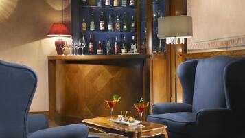 Uma área de bar com dois poltronas azuis, uma pequena mesa de madeira com bebidas, e um balcão repleto de garrafas de licor.