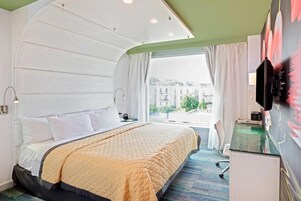 Quarto de hotel com cama, escrivaninha, cadeira, televisão e janela com vista para edifícios.