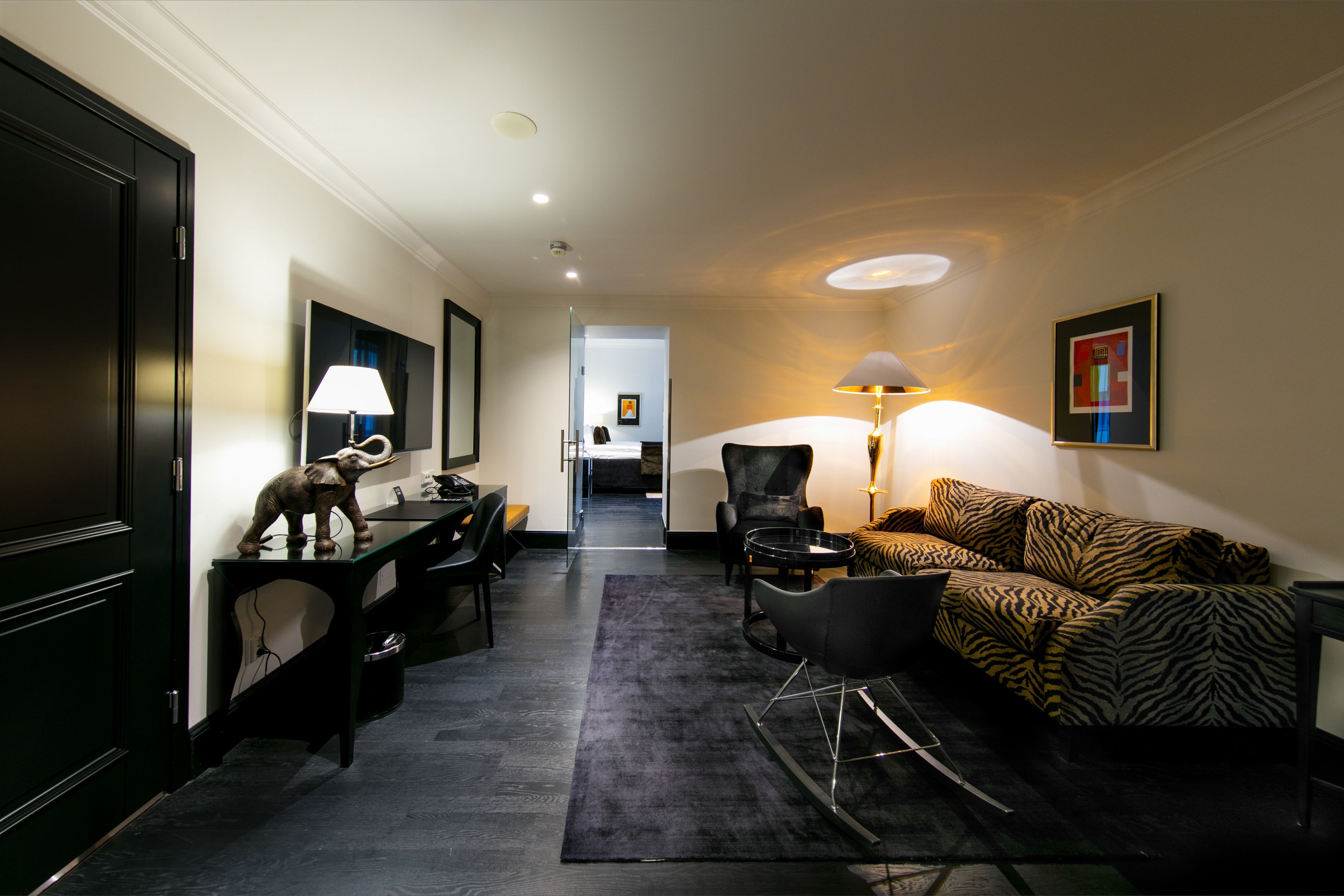 Quarto de hotel com escrivaninha de madeira escura, sofá estampado com padrão de zebra, poltrona preta e tapete preto.