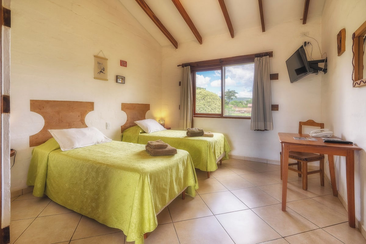 Quarto com duas camas, uma escrivaninha, uma cadeira, televisão e janela com cortinas.